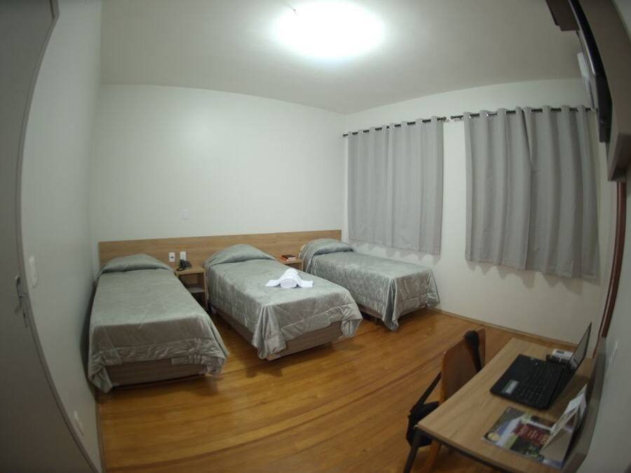 Quarto com duas camas de solteiro, uma escrivaninha de madeira com um laptop e uma janela com cortinas.