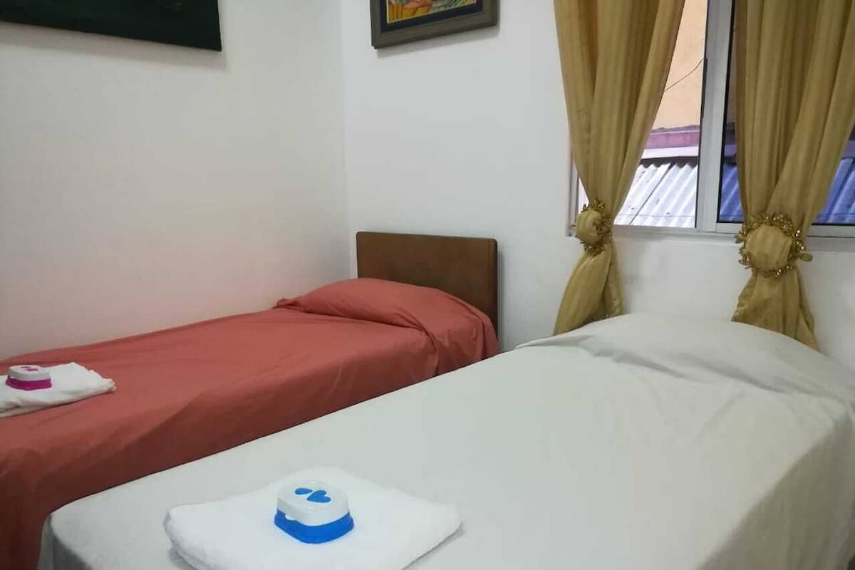Quarto de hotel com duas camas, uma com colcha vermelha e a outra com colcha branca. Há uma janela com cortinas bege, um quadro emoldurado na parede e uma toalha branca com logótipo azul na cama.
