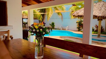 Vista da piscina com mesa de madeira, um vaso de flores e um guarda-sol de palha.