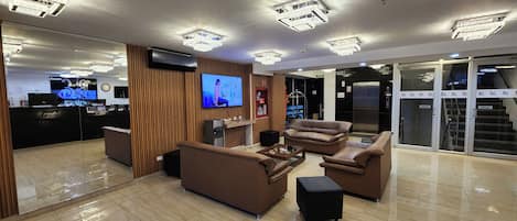 Um lobby moderno com balcão de recepção, TV de tela plana, sofás de couro e um elevador de vidro.