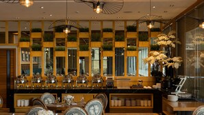 Um restaurante com um balcão comprido, várias mesas e cadeiras. Há plantas na parede e uma grande janela no lado direito.