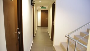Um corredor de hotel com uma porta de madeira à esquerda, uma placa indicando o caminho para a cozinha e uma escada à direita.