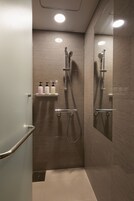 Um chuveiro moderno com um chuveiro manual e torneira fixada na parede, um pequeno armário com produtos de higiene e um espelho refletindo o espaço do chuveiro.