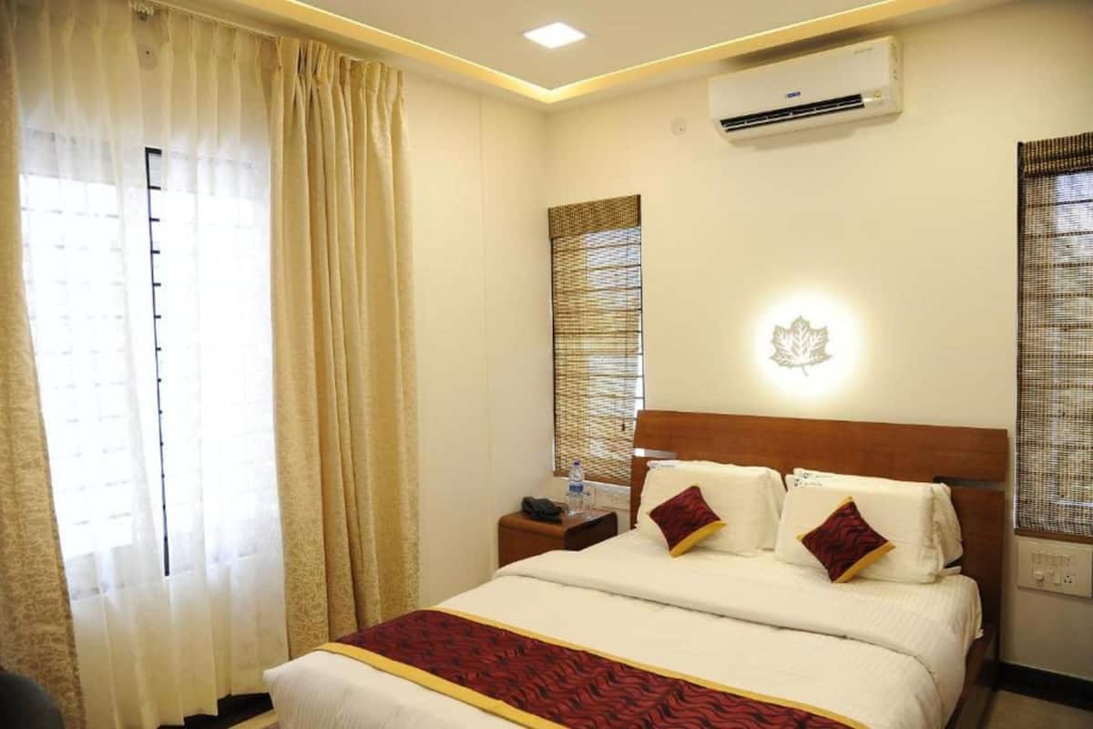 Quarto de hotel com cama, cortinas, unidade de ar condicionado, mesa de cabeceira com telefone e garrafa de água, e luminária na parede.