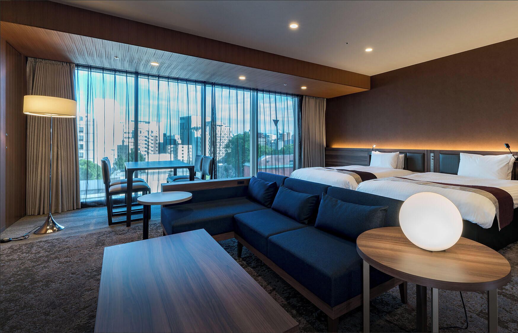 Quarto de hotel moderno com uma cama grande, um sofá, uma mesa de jantar e vista para a cidade.