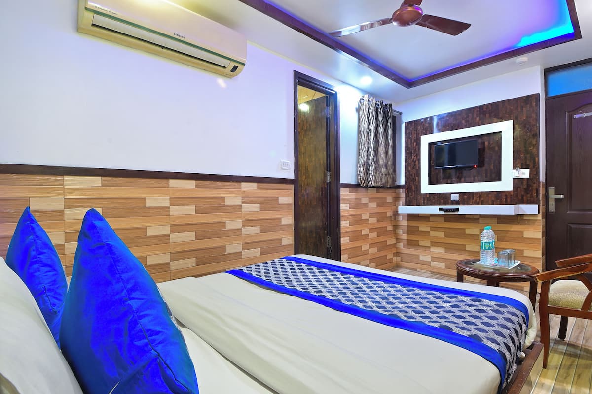 Quarto de hotel com cama, televisão fixada na parede, ventilador de teto e piso de madeira.