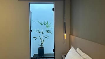 Quarto com cama, janela e vista para plantas, e mesa de cabeceira.