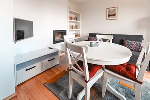 Uma sala de estar moderna com uma mesa de jantar branca, cadeiras brancas, um sofá cinza com almofadas vermelhas, uma estante e uma televisão de tela plana.