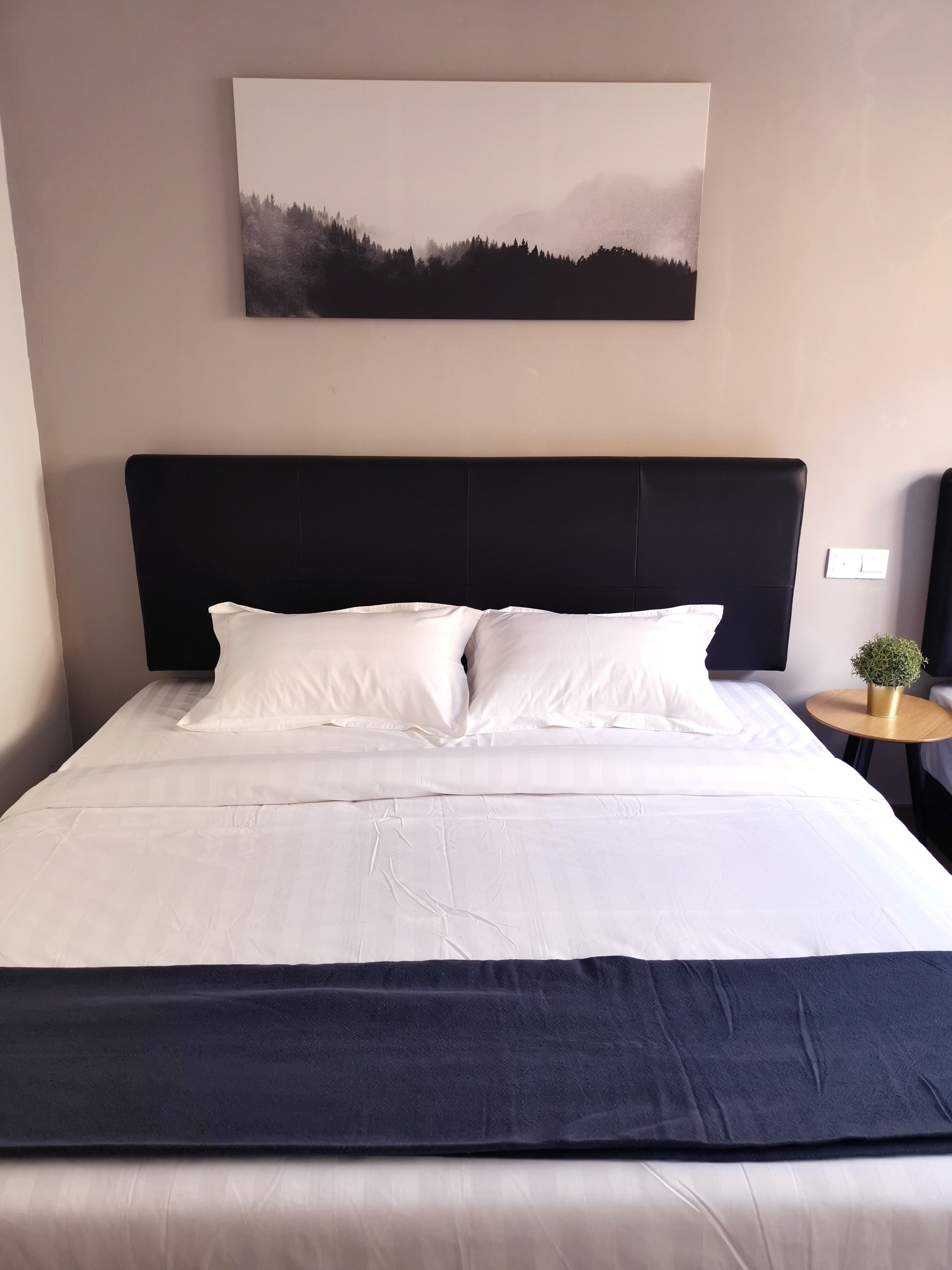 Uma cama bem arrumada com roupa de cama branca e cabeceira escura, uma pequena mesa redonda com uma planta e um quadro de uma floresta na parede.