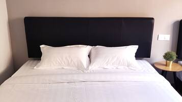 Uma cama bem arrumada com roupa de cama branca e cabeceira escura, uma pequena mesa redonda com uma planta e um quadro de uma floresta na parede.