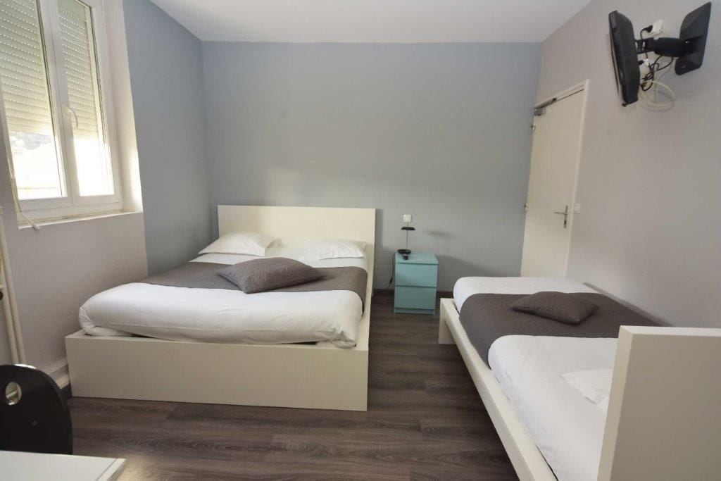 Quarto com duas camas, uma mesa de cabeceira, uma luminária e uma televisão fixada na parede.