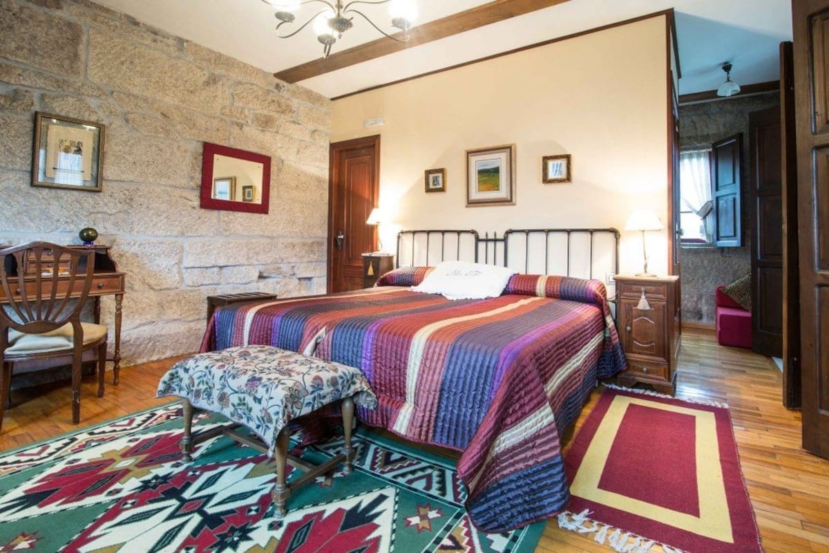 Quarto com parede de pedra, cama de madeira, escrivaninha e cadeira.