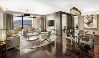 Quarto de hotel moderno com uma janela grande que oferece vista para as montanhas, uma área de estar aconchegante com sofá e poltronas, uma mesa de jantar com cadeiras e uma TV de tela plana fixada na parede.