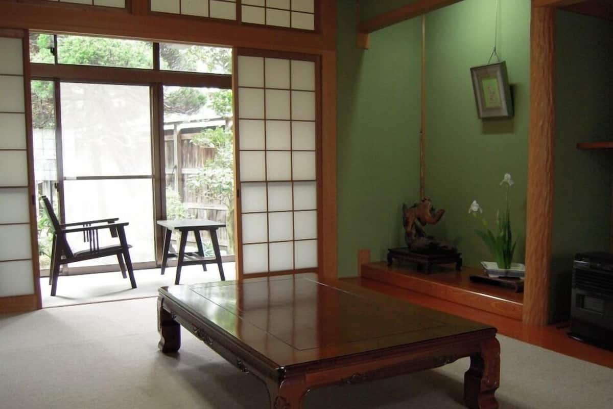 Um quarto japonês tradicional com tatames, uma mesa de madeira baixa, uma porta deslizante que dá para um jardim e uma parede verde com uma moldura.