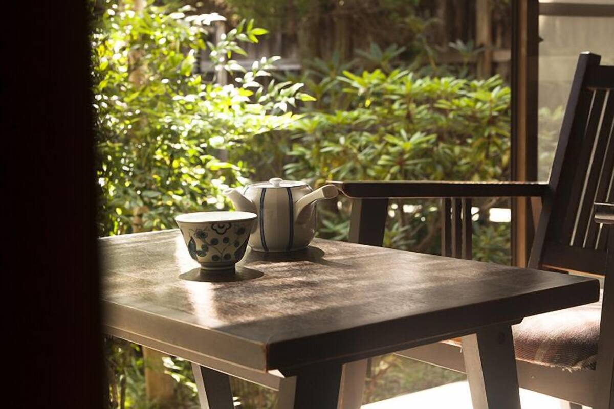 Uma mesa com um bule e uma xícara, uma cadeira e vista para o verde lá fora.