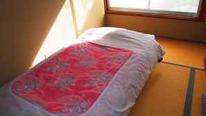 Cama de solteiro com colchão branco e edredom rosa floral em um quarto com piso de tatame e janela.