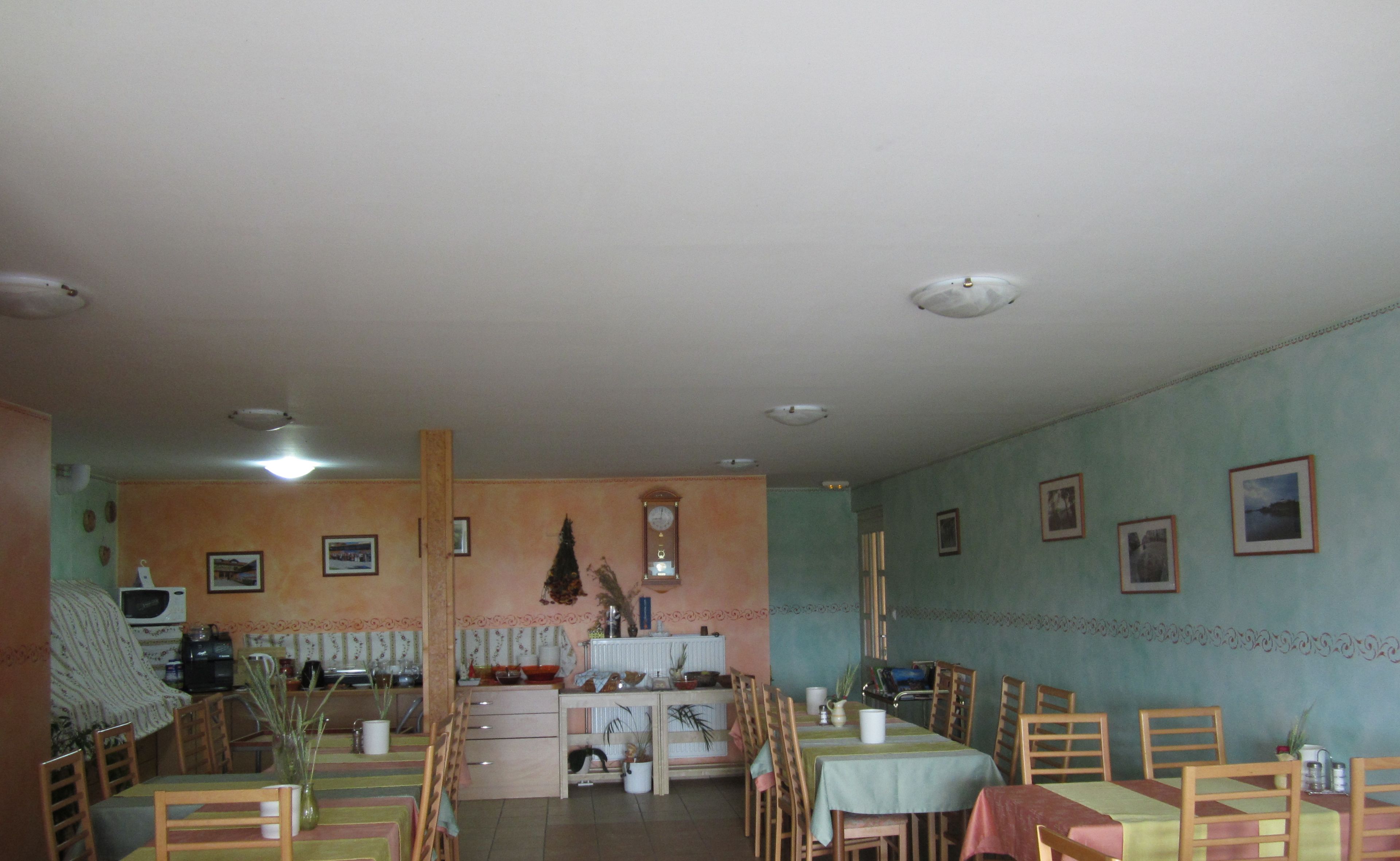 Uma sala de jantar com cozinha ao fundo, cadeiras de madeira, uma mesa com toalha e um relógio na parede.