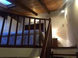 Um quarto com teto de madeira, uma cama com roupa de cama branca, uma mesa de cabeceira com abajur e uma janela com persianas.