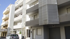 Um edifício residencial com vários andares, varandas, um carro estacionado em frente e uma porta com persiana fechada.