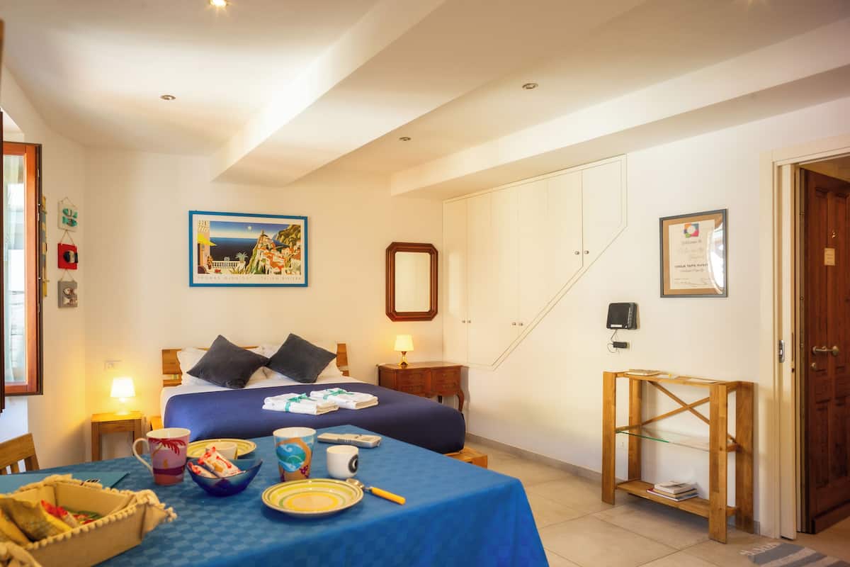 Quarto de hotel com cama, mesa de cabeceira, luminária e uma pequena mesinha de madeira.