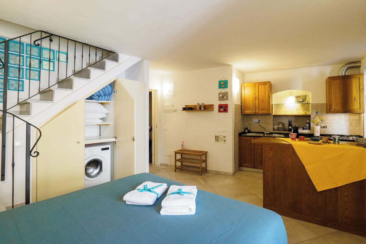 Quarto de hotel compacto com cama, máquina de lavar, uma mesinha e cozinha equipada com armários de madeira e pia.