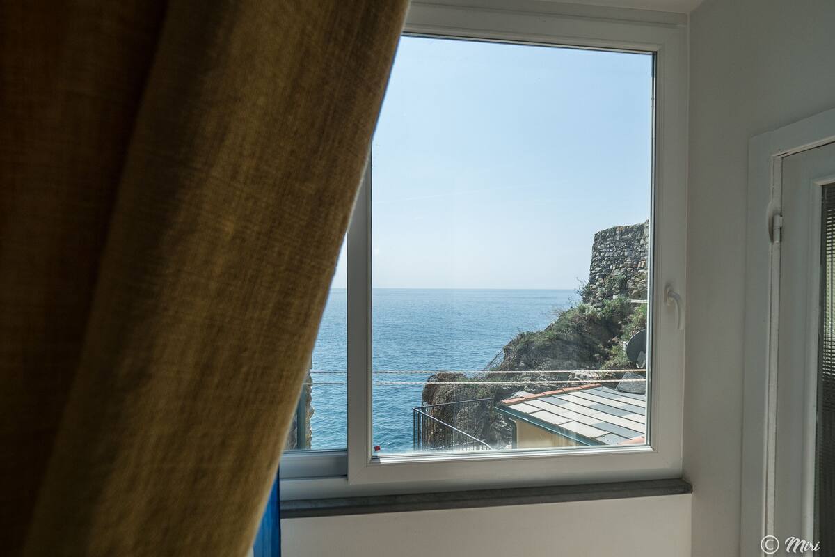 Quarto com janela que oferece vista para a costa, incluindo um penhasco rochoso e o mar.