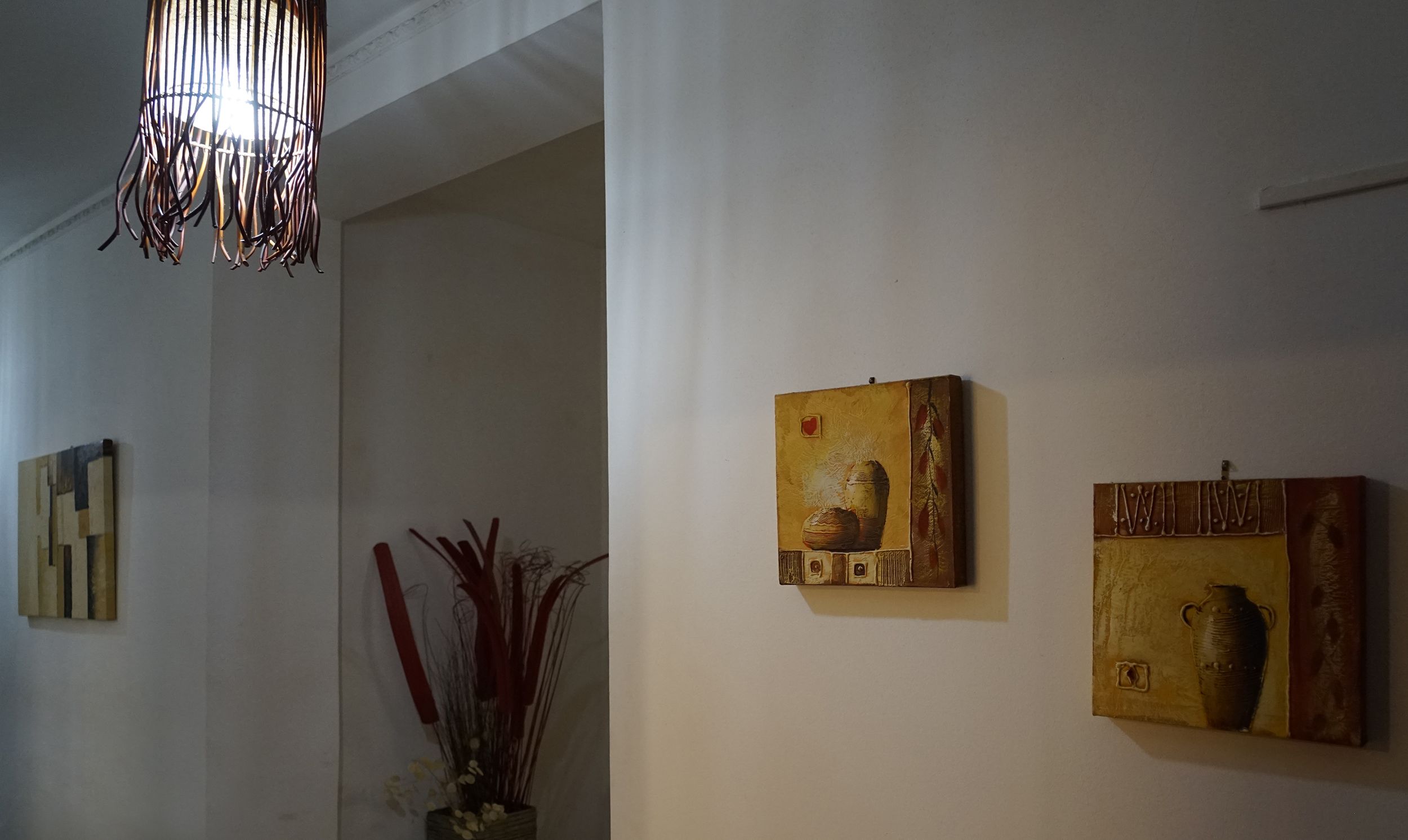 Um corredor com obras de arte nas paredes, uma luminária pendente e um vaso de planta.