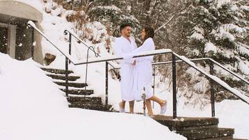 Um casal vestido com túnicas brancas em uma escadaria coberta de neve, com guarda-corpos de metal, cercado por árvores carregadas de neve.