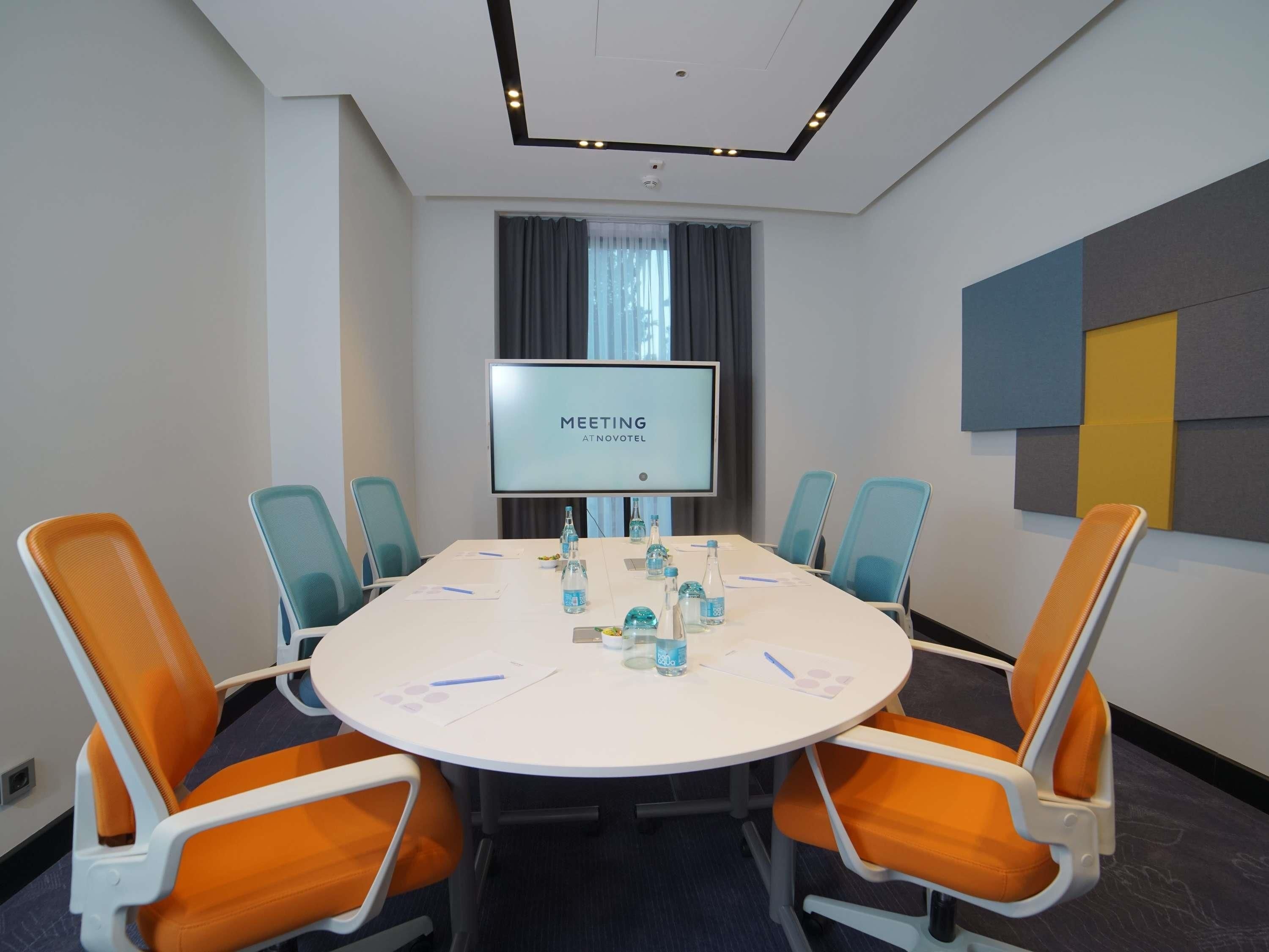 Uma sala de conferências com uma mesa redonda, seis cadeiras laranja, uma tela grande exibindo 'MEETING ATNOVOTEL' e um design de teto moderno.