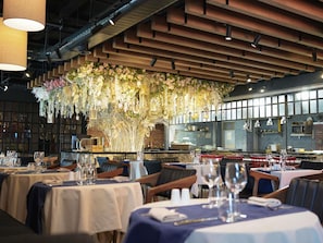 Restaurante com mesas preparadas para refeições, cozinha ao fundo e um arranjo floral decorativo pendurado.