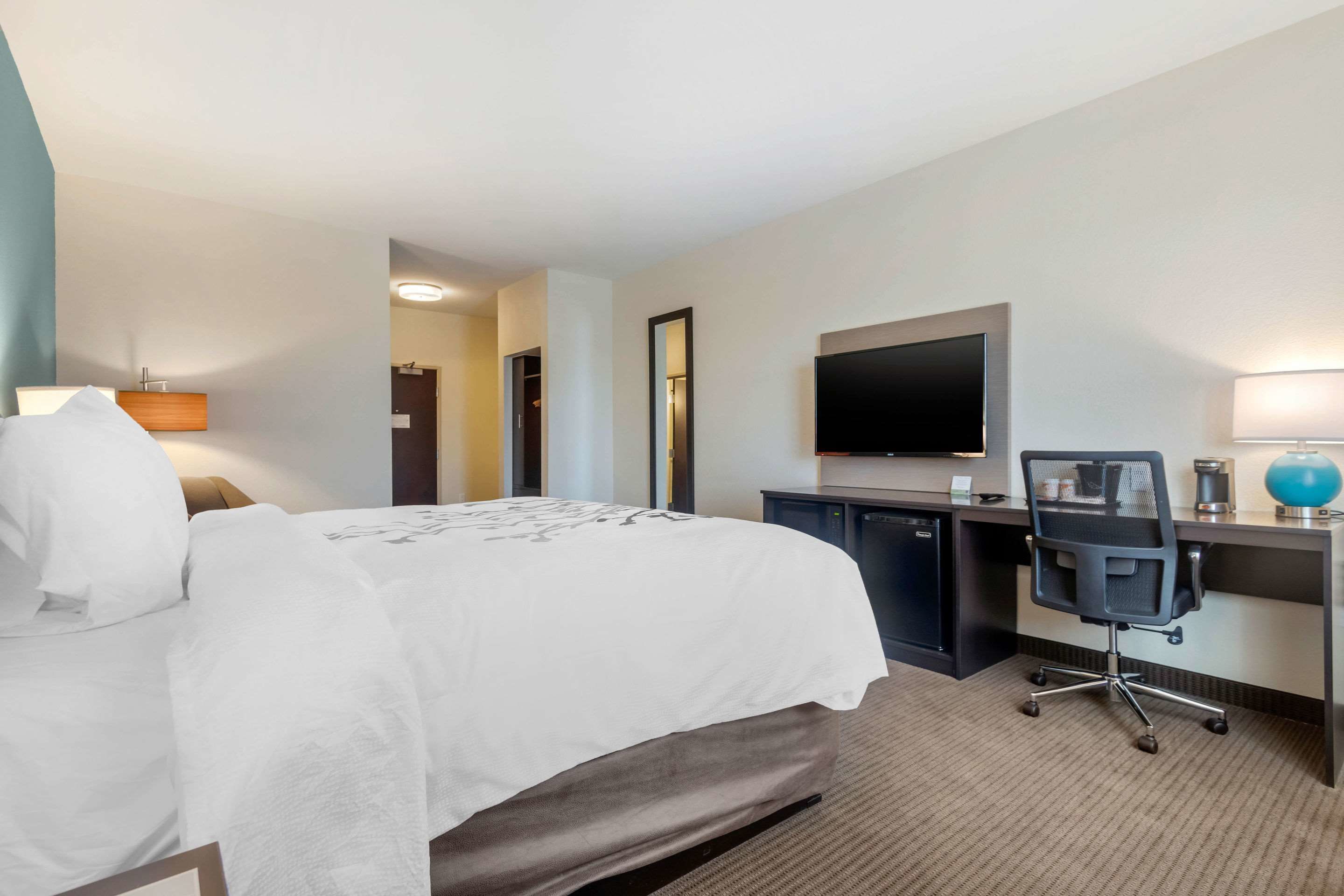 Quarto de hotel com cama, escrivaninha, cadeira, televisão e luminária.