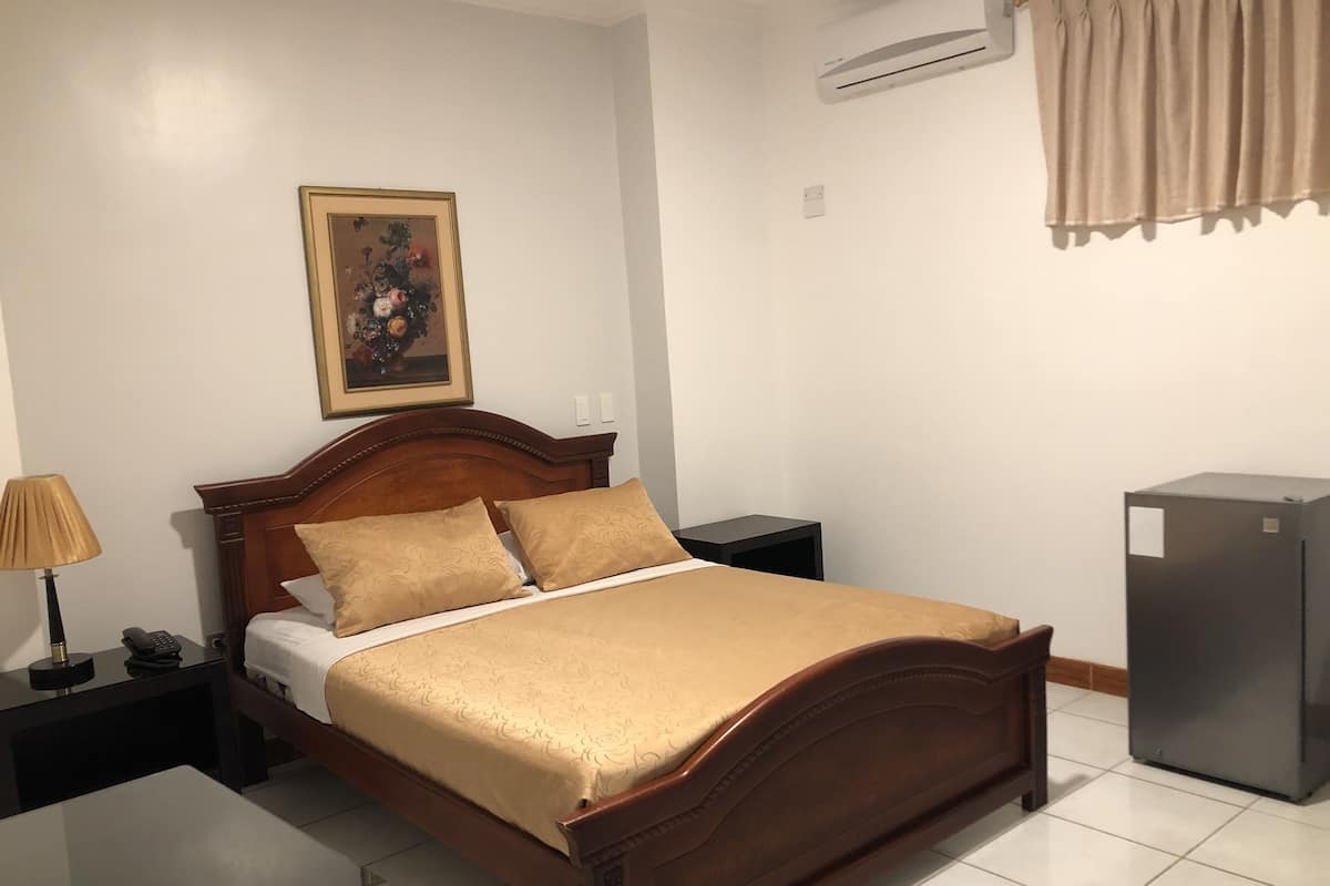 Quarto bem arrumado com cama de madeira, mesa de cabeceira com luminária, uma pequena mesa, um refrigerador e um quadro na parede.