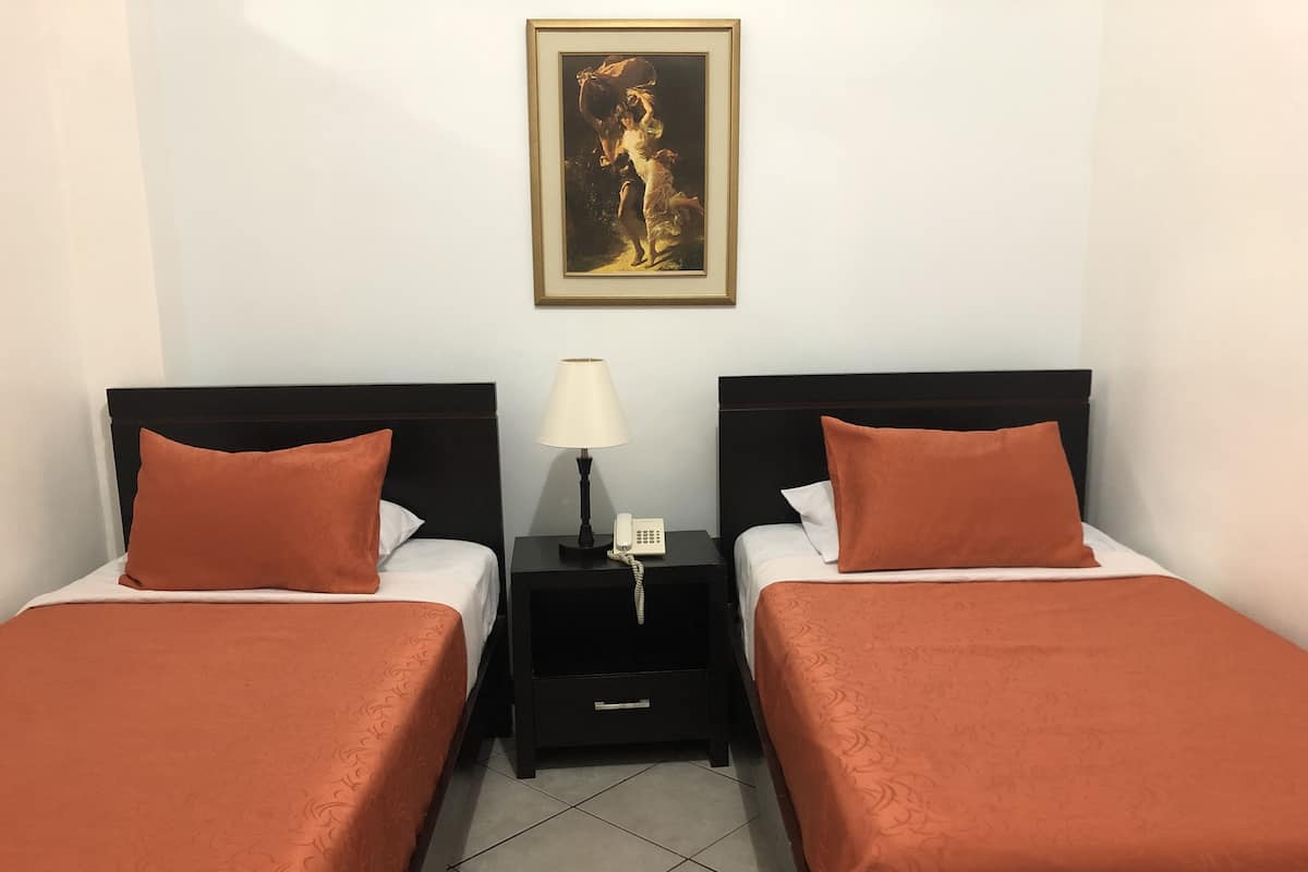Quarto de hotel com duas camas, cada uma com colcha laranja e almofadas a combinar, mesa de cabeceira com abajur e telefone, e quadro pendurado na parede.