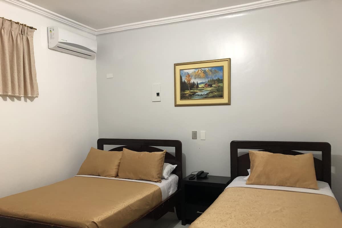 Quarto com duas camas de solteiro, roupa de cama bege, um quadro na parede, ar condicionado e cortina.