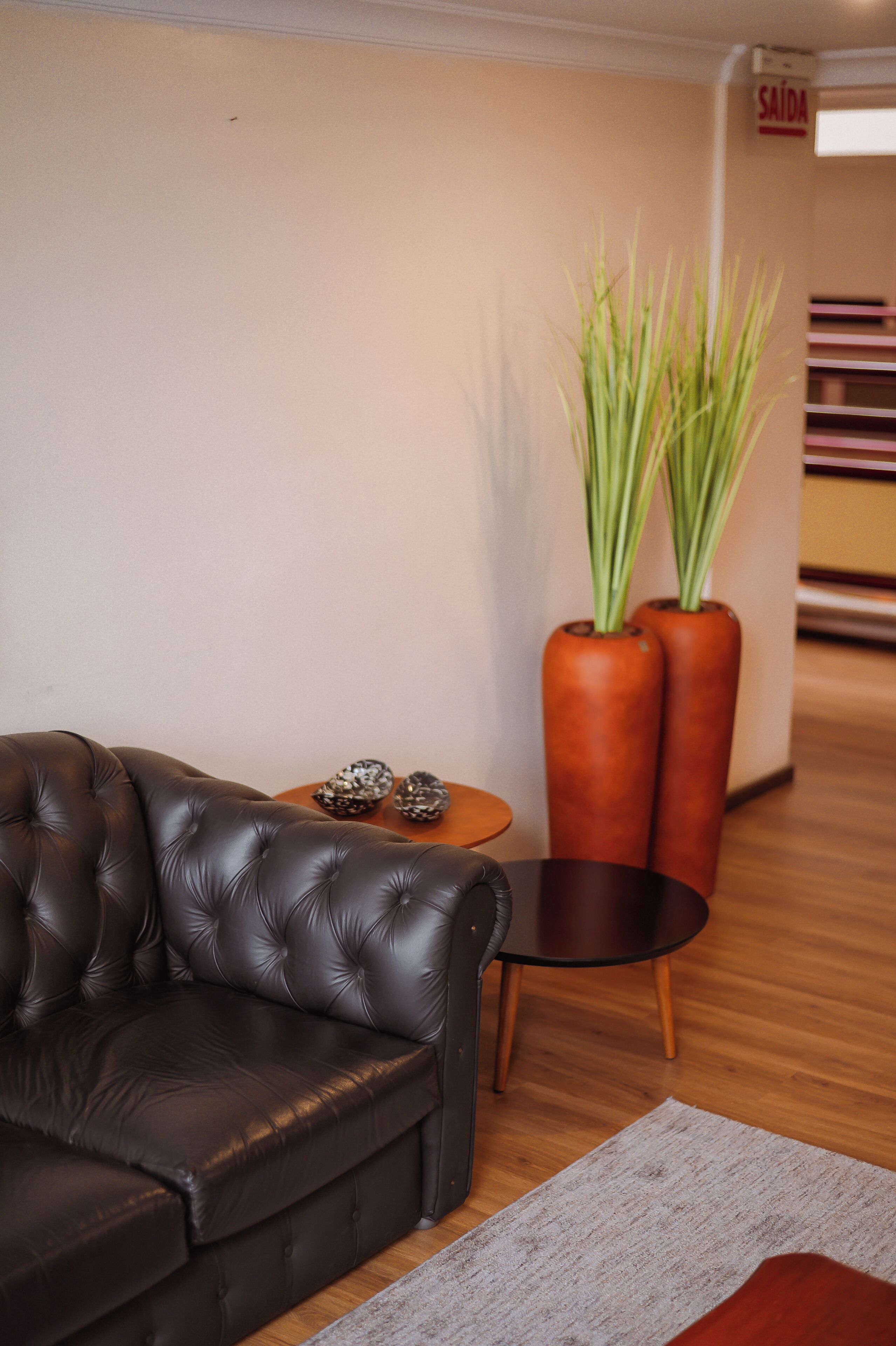 Uma sala de estar moderna com um sofá de couro escuro, uma pequena mesa lateral de madeira e dois vasos altos laranja com plantas verdes.