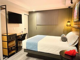Quarto de hotel com uma cama, uma escrivaninha, uma cadeira, uma TV e uma mala vermelha sobre uma mesinha.
