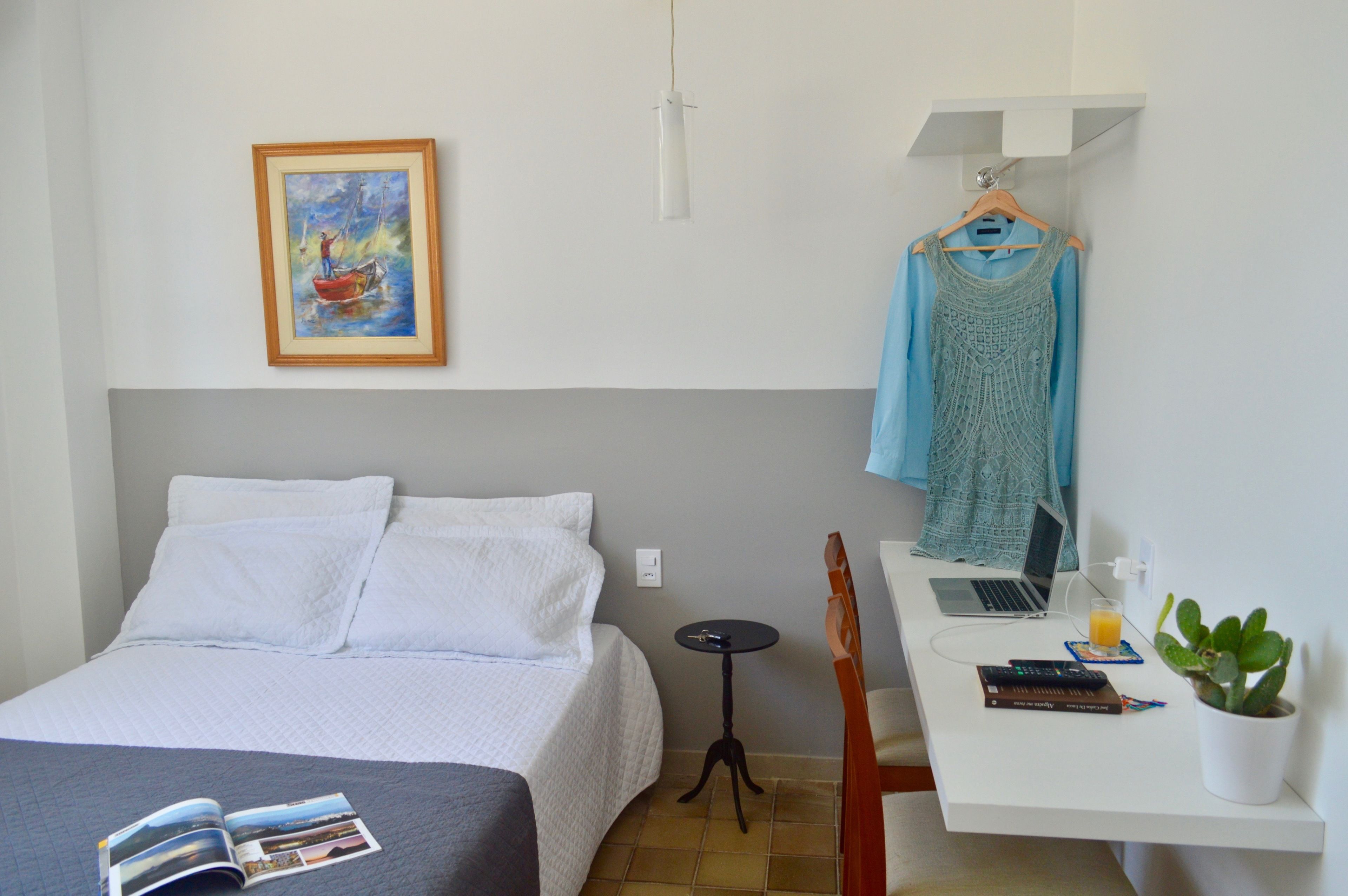Quarto bem arrumado com uma cama, uma escrivaninha com um laptop, um vestido pendurado, um quadro emoldurado e um vaso de planta.
