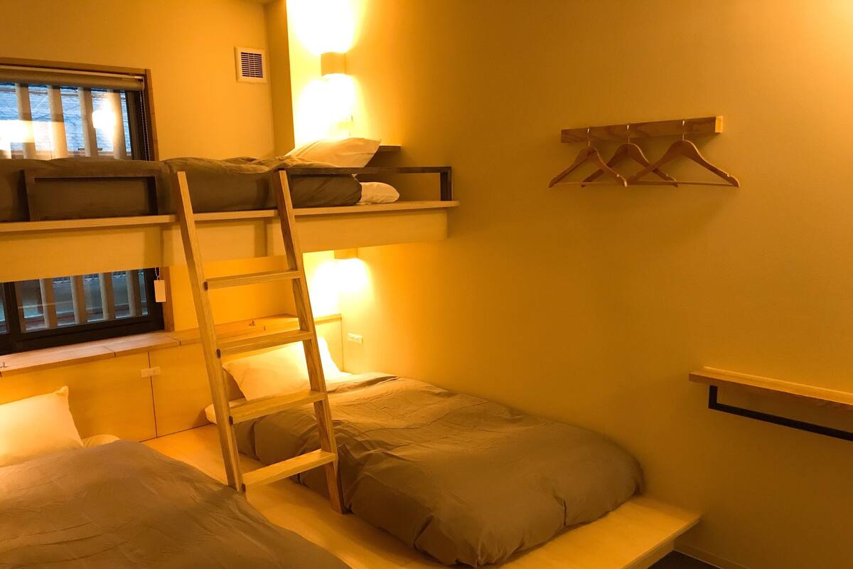 Quarto compacto com beliches, escada e janela com persianas.