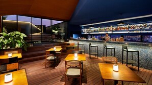 Uma área de bar moderna com mesas e cadeiras de madeira, balcão com diversos tipos de garrafas, e vista da cidade ao entardecer.