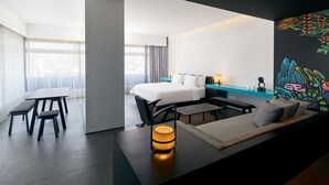 Quarto de hotel moderno com cama, sofá, mesa de jantar com bancos e uma pequena mesa com abajur.