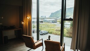 Quarto de hotel com duas cadeiras, uma mesa e uma janela grande com vista para um estacionamento e edifícios.