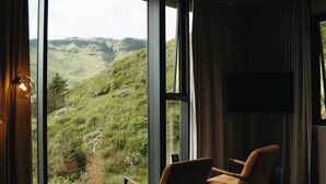 Quarto com grandes janelas com vista para um vale verde. Há duas poltronas de couro e uma televisão de tela plana fixada na parede.