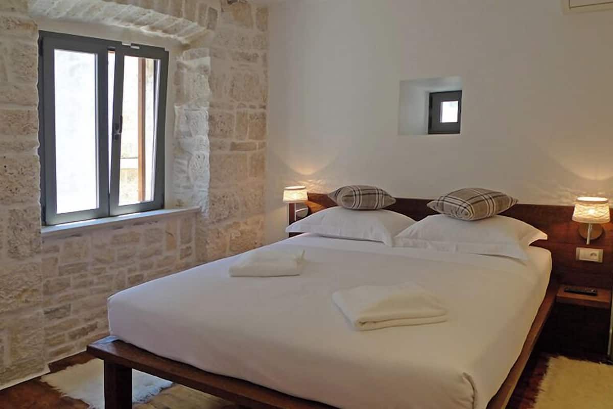 Quarto com parede de pedra, janela, cama com lençóis brancos, duas luminárias de cabeceira e um pequeno espelho na parede.