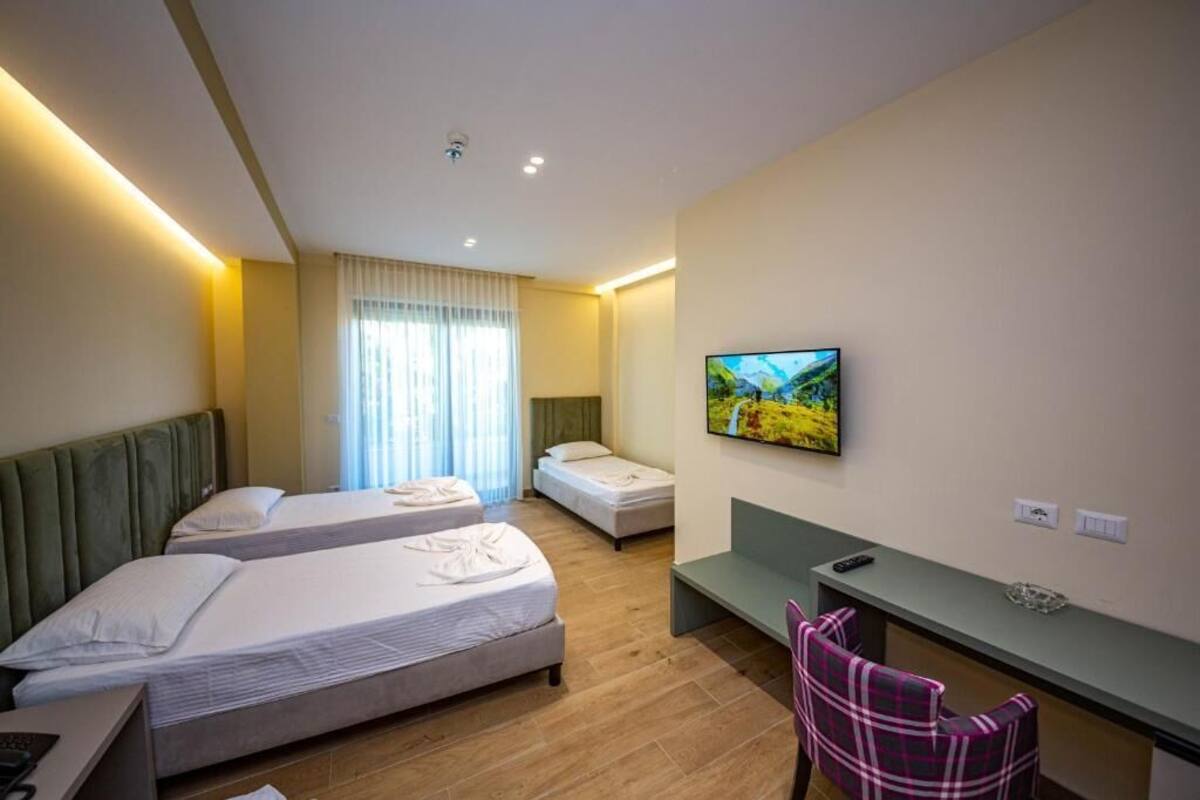 Quarto de hotel com duas camas, uma mesa, uma cadeira, uma TV e uma janela com cortinas.