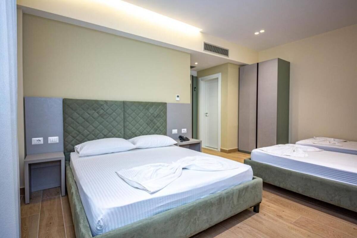 Quarto de hotel com duas camas, cabeceira verde, mesa de cabeceira e guarda-roupa.