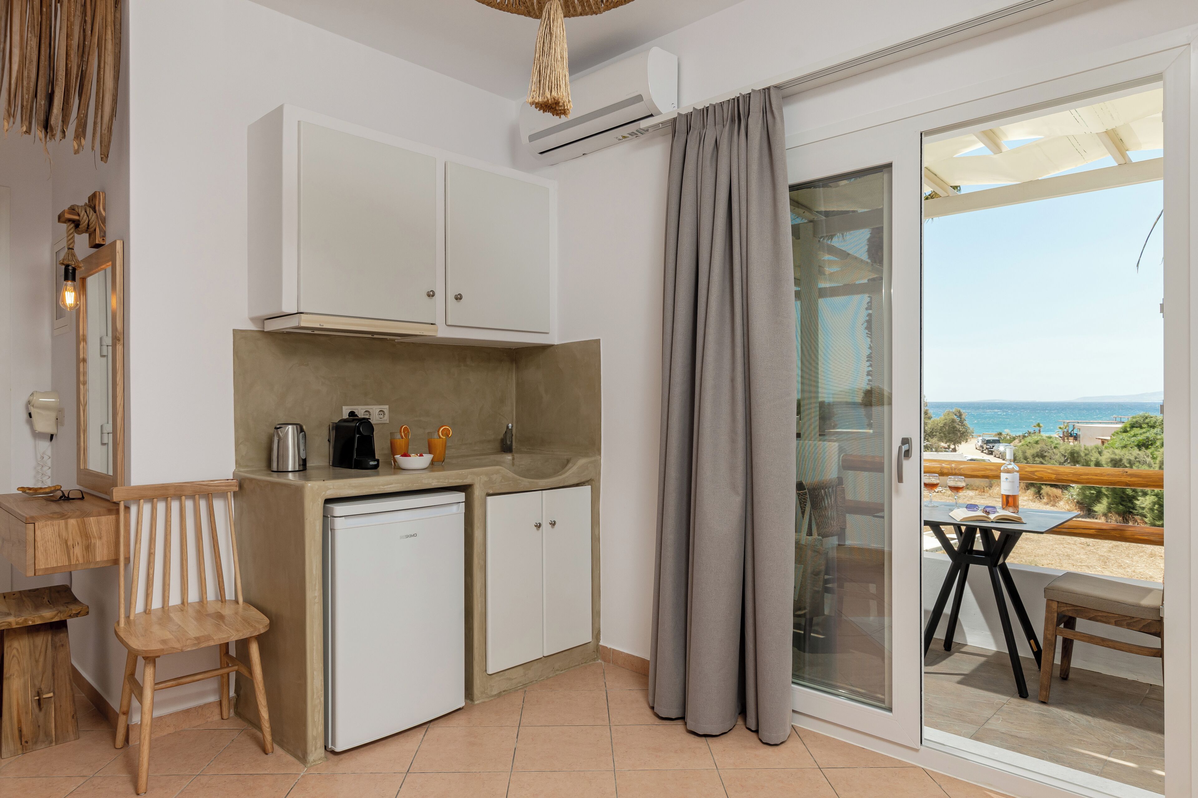 Cozinha compacta com máquina de lavar louça, cafeteira e chaleira na bancada. Uma cadeira de madeira e uma mesa de jantar com um livro e uma garrafa sobre ela. O quarto tem uma porta de vidro deslizante que dá para uma varanda com vista para o mar.