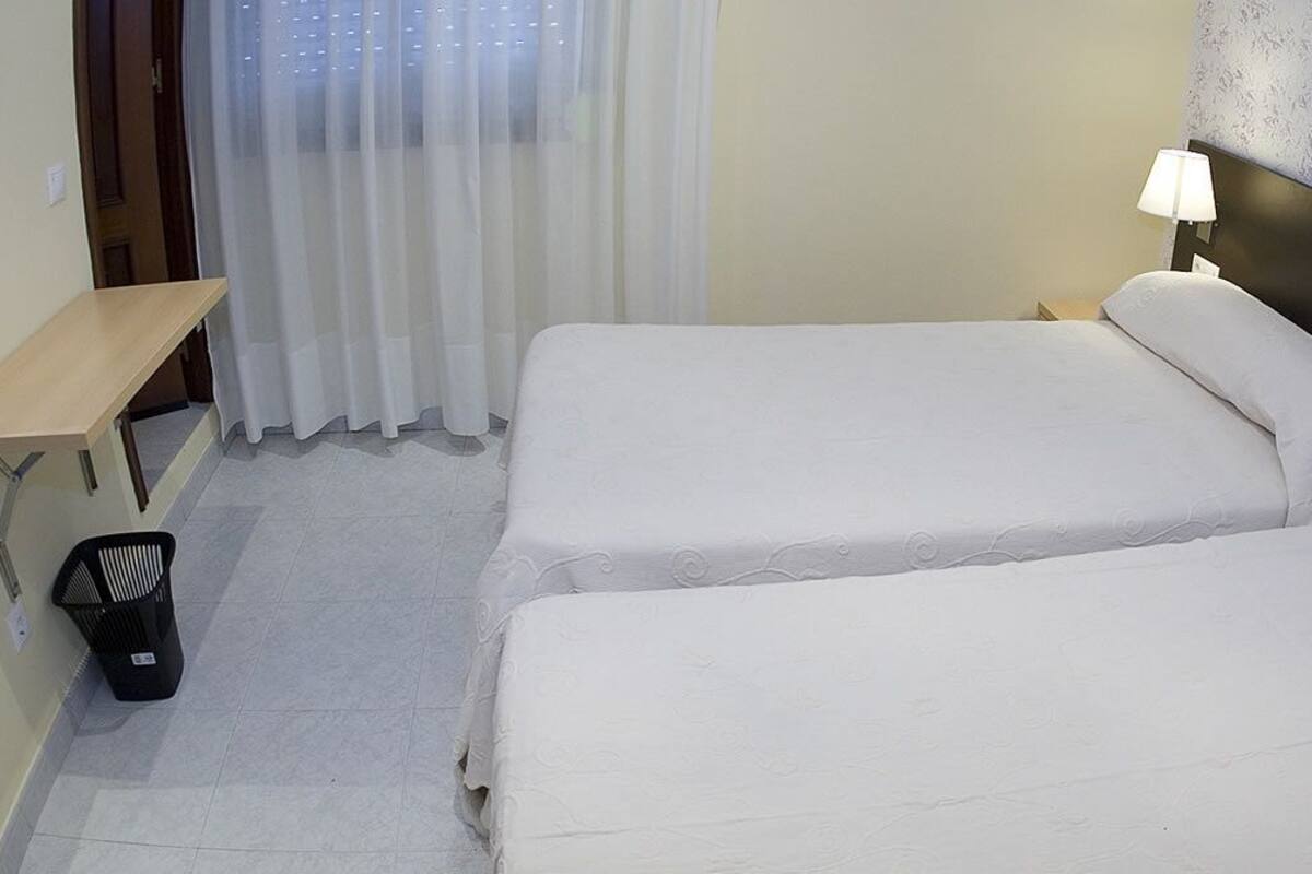 Quarto de hotel com duas camas, uma escrivaninha, uma cadeira e uma janela com cortinas.