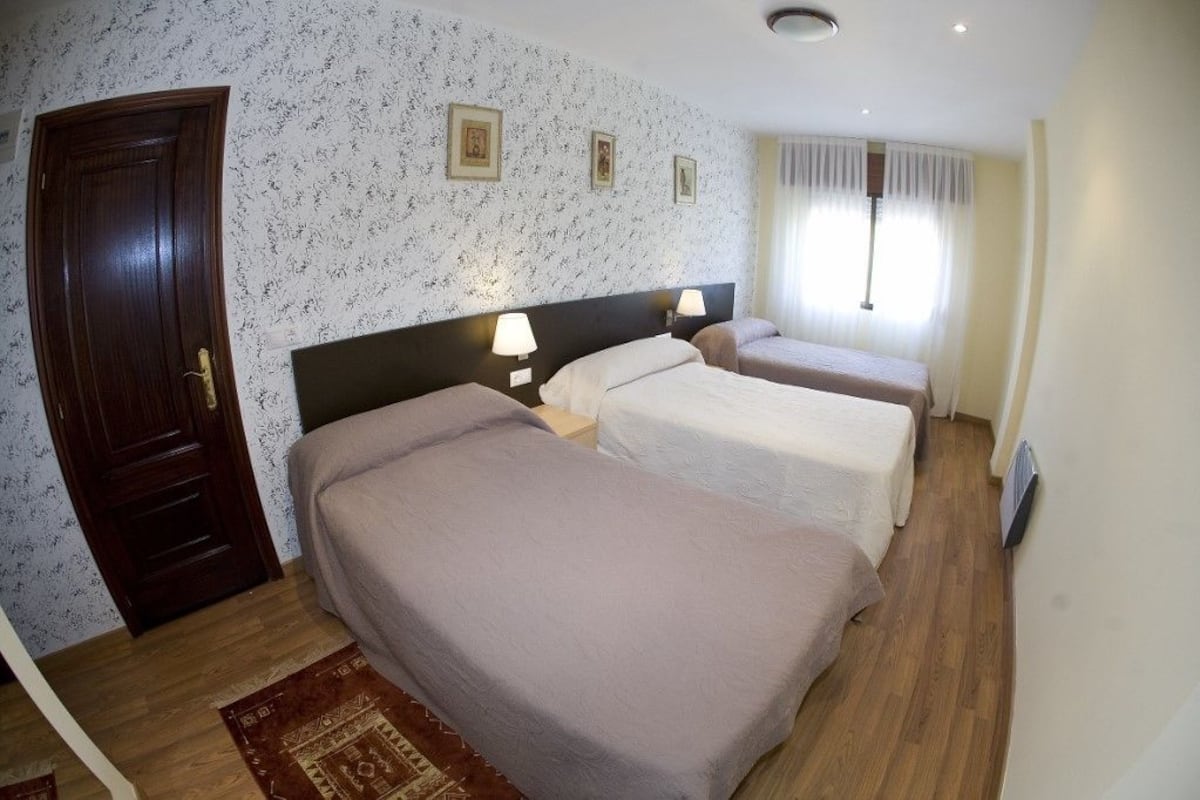 Quarto de hotel com duas camas, porta de madeira, papel de parede estampado e janela com cortinas.