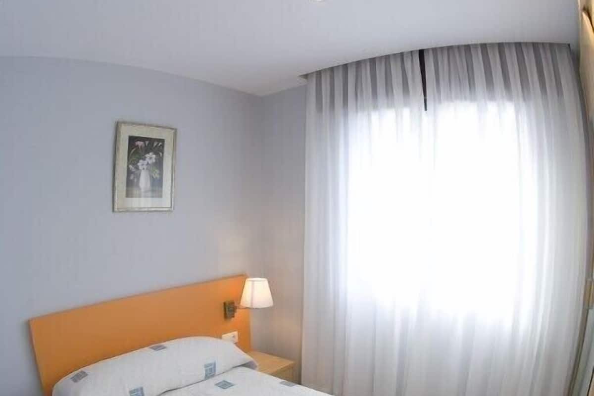 Quarto de hotel com uma cama de solteiro, mesa de cabeceira com abajur, quadro na parede e janela com cortinas.