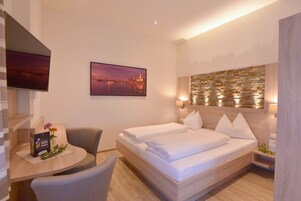 Quarto de hotel com cama, escrivaninha, cadeira e televisão na parede.
